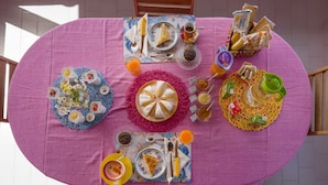 Mesa posta com diversos itens de café da manhã, incluindo doces, frutas e bebidas, sobre uma toalha de mesa rosa.