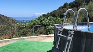 Piscina com escada, rodeada por relva sintética verde e vista para uma paisagem ondulada com um corpo de água ao longe.