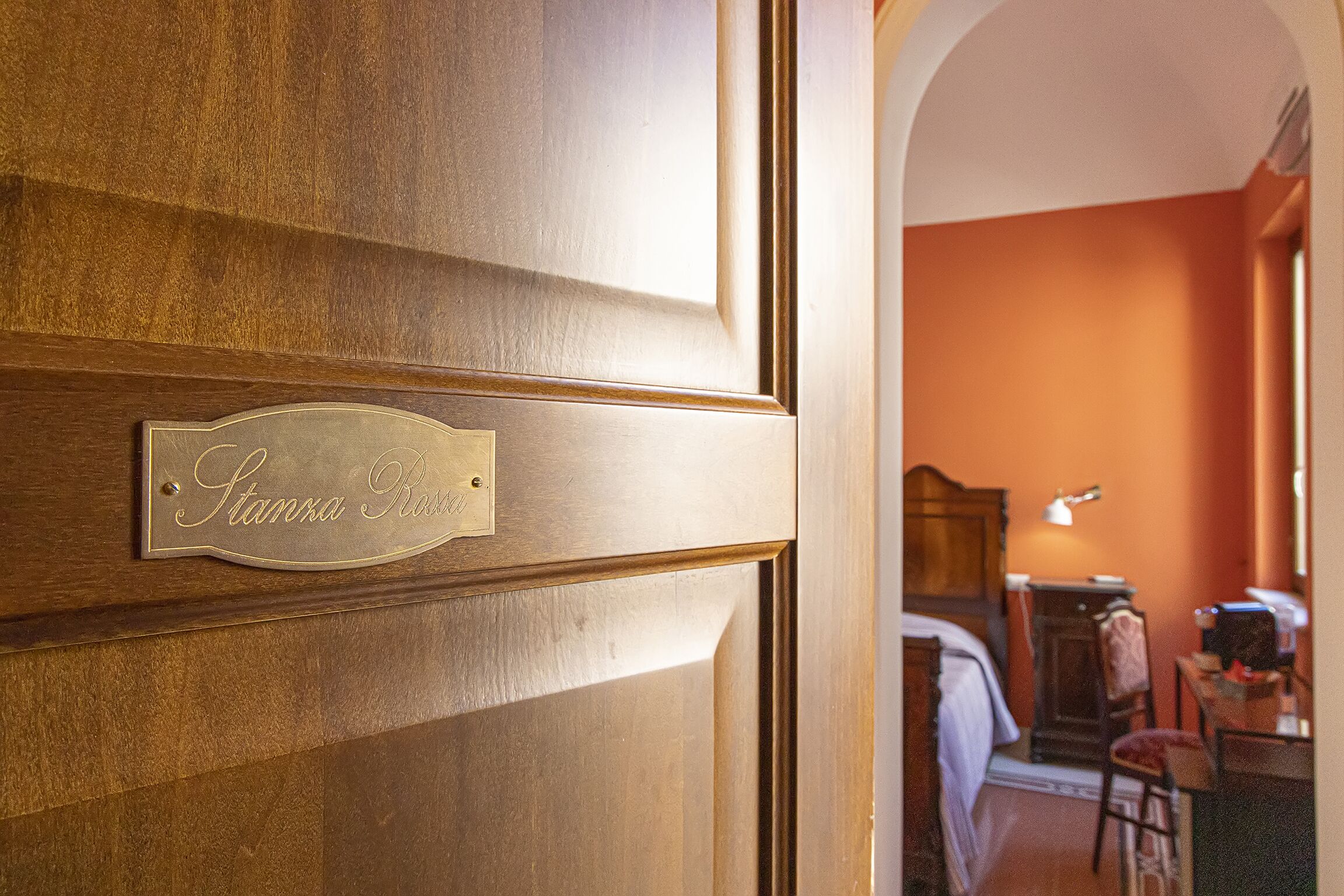 Quarto de hotel com porta de madeira, placa com nome, cama, escrivaninha, cadeira e luminária.