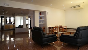 Um hall amplo com uma mesa de recepção, dois sofás de couro preto, uma área de refeições com cadeiras de madeira e piso de mármore.
