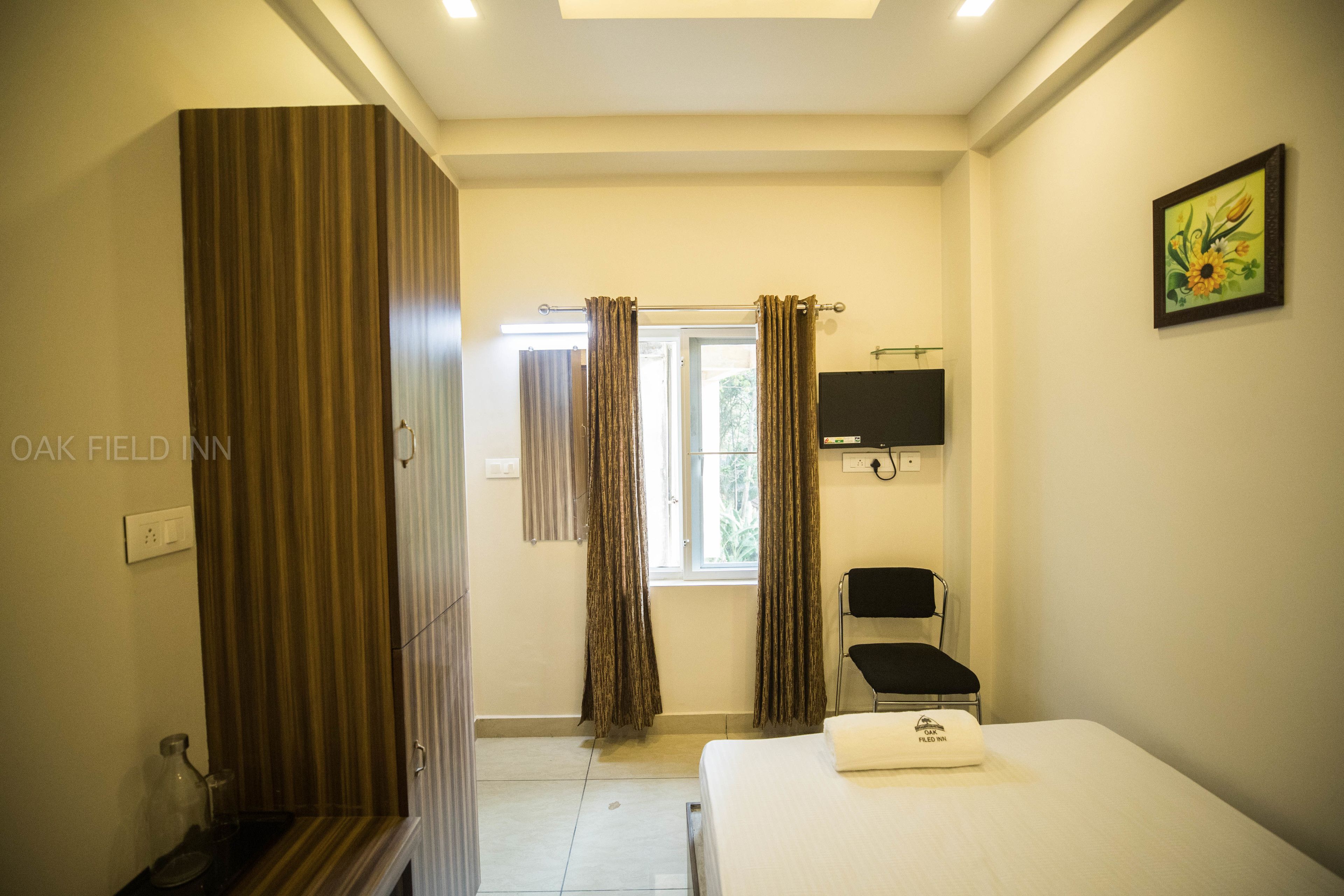 Quarto de hotel com cama, uma cadeira, televisão fixada na parede e janela com cortinas.