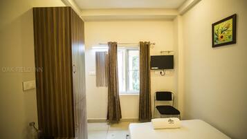 Quarto de hotel com cama, uma cadeira, televisão fixada na parede e janela com cortinas.