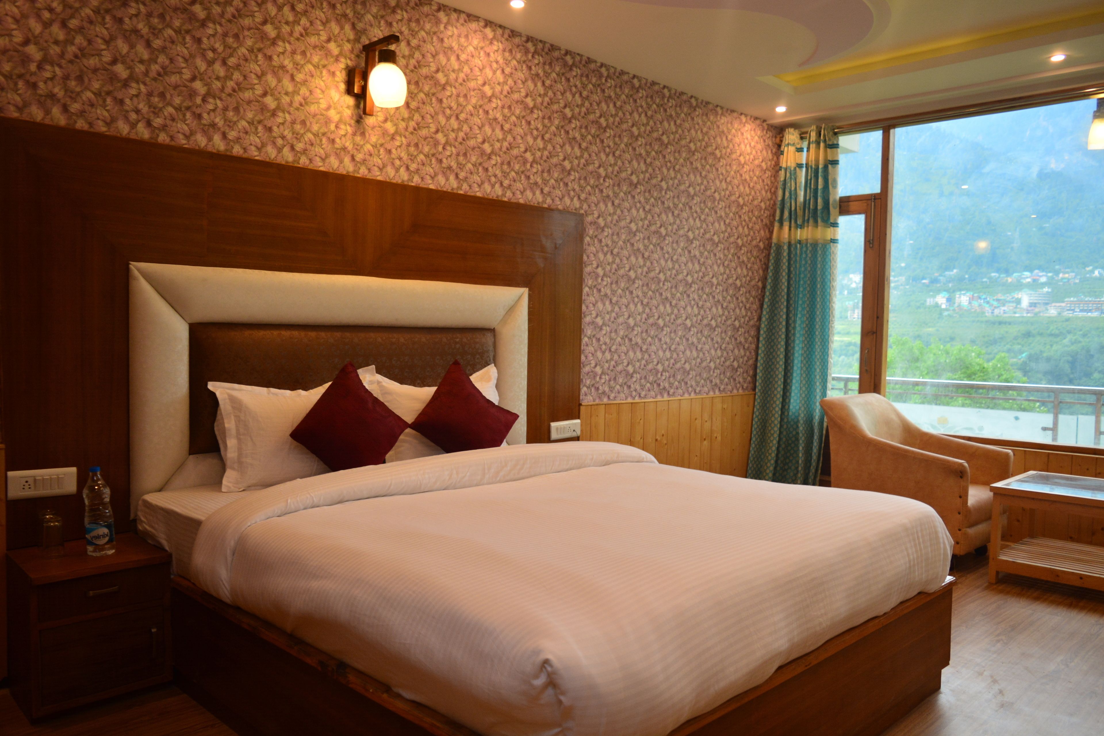 Quarto de hotel com uma cama grande, cabeceira de madeira, mesa de cabeceira, uma cadeira e vista para o exterior.