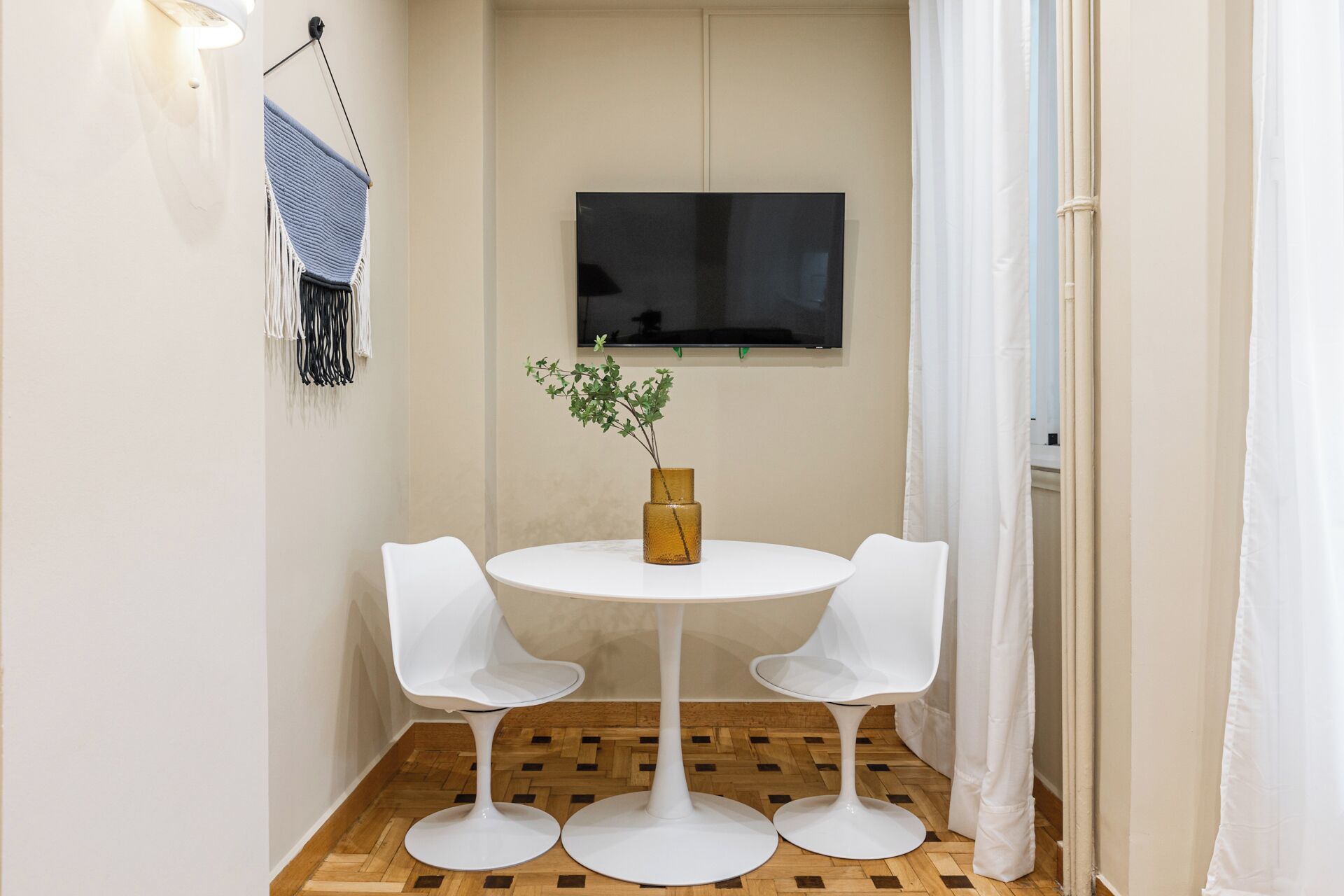 Um quarto pequeno e bem iluminado com uma mesa redonda e duas cadeiras brancas, uma pequena janela com uma televisão e uma planta em vaso.