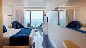 Quarto de hotel moderno com cama, escrivaninha, cadeira e uma grande janela com vista para o oceano.