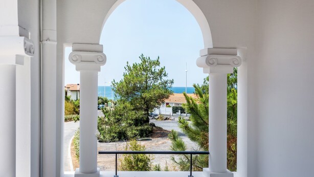 Vista através de uma porta em arco com colunas brancas, dando para uma área residencial com árvores e um edifício com telhado em cerâmica.