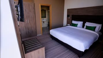 Quarto de hotel com uma cama grande, televisão fixada na parede, um armário de madeira e um banheiro visível através de uma porta aberta.