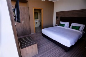 Quarto de hotel com uma cama grande, televisão fixada na parede, um armário de madeira e um banheiro visível através de uma porta aberta.
