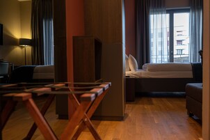 Quarto de hotel com piso de madeira, sofá, cama, escrivaninha com luminária e vista para a cidade pela janela.