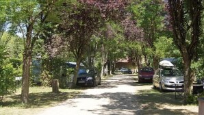 Uma área de estacionamento com carros estacionados sob árvores, um caminho de cascalho e um edifício ao fundo.