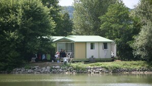 Uma pequena cabana térrea com telhado verde, localizada à beira de um lago. Há áreas de estar ao ar livre com cadeiras e mesas, e uma pessoa pode ser vista dentro da cabana.