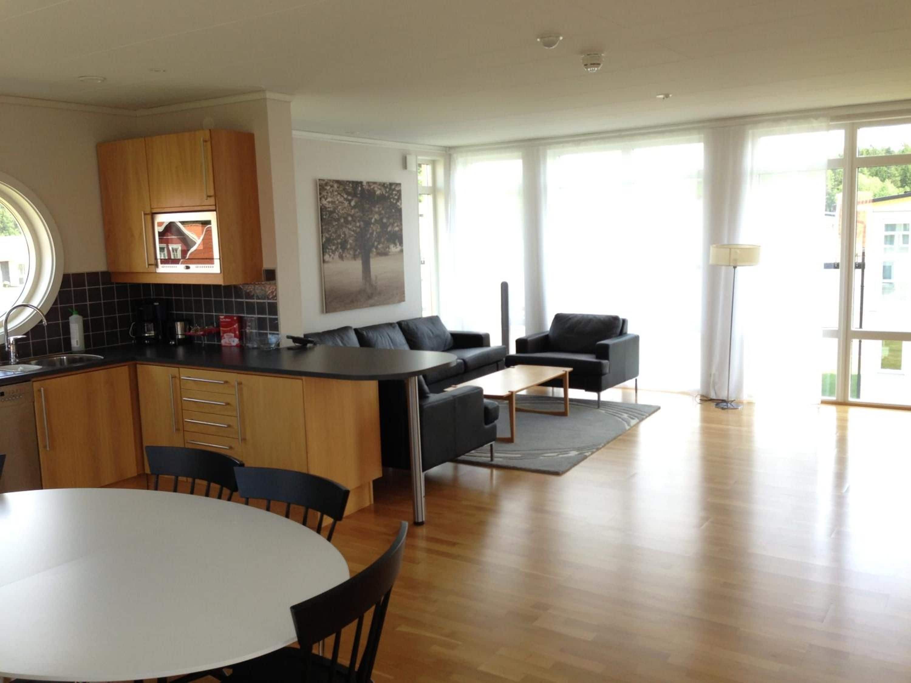 Cozinha moderna com ilha central, área de jantar com mesa e cadeiras redondas, sofá preto e uma janela grande que oferece iluminação natural.