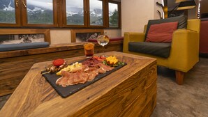 Uma mesa de madeira com uma tábua de frios, um copo de vinho tinto e vista para montanhas através de grandes janelas.