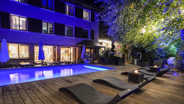 Uma área de piscina à noite, com espreguiçadeiras, guarda-sóis e um edifício com janelas iluminadas.