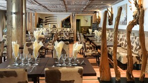 Restaurante com vigas de madeira no teto, cadeiras estofadas em couro de vaca e mesas postas com copos de vinho e guardanapos.