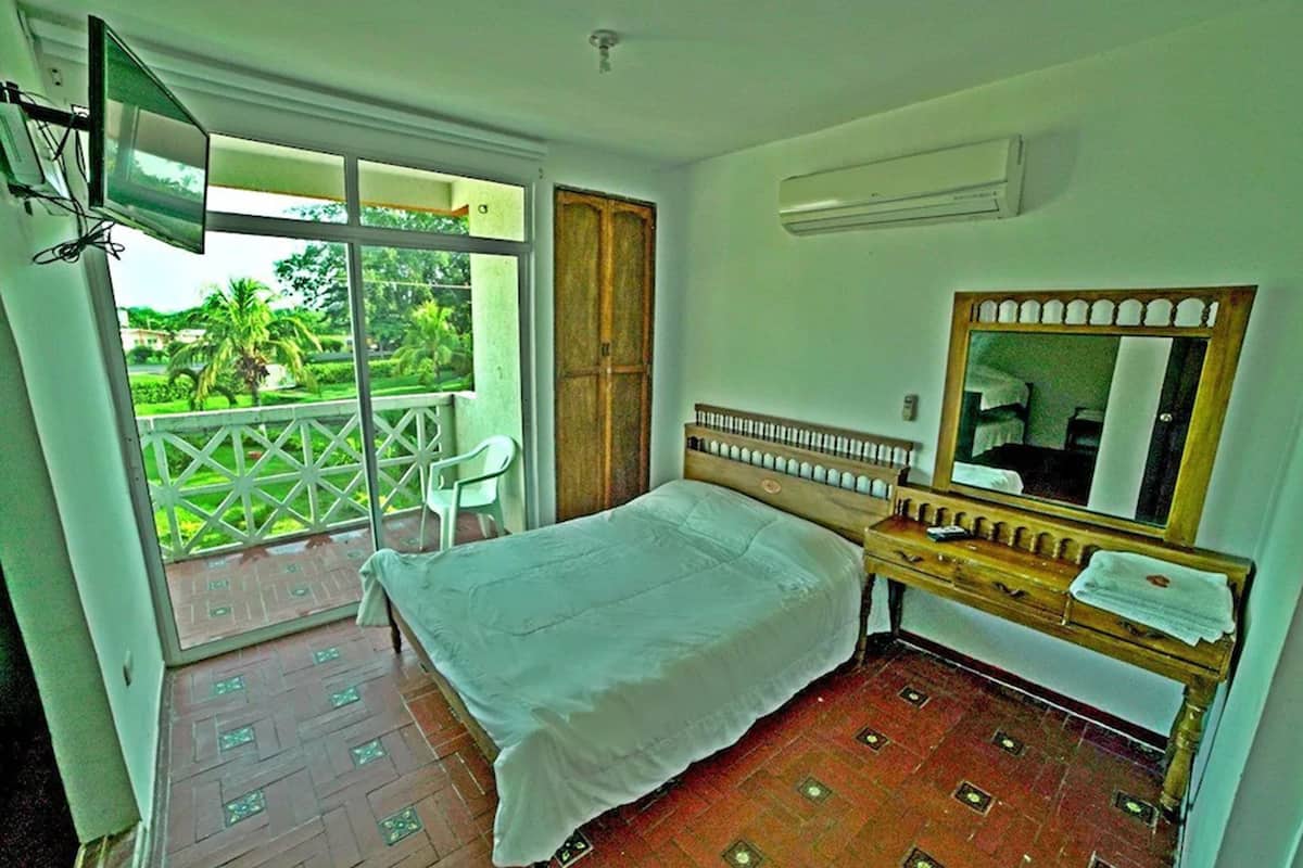 Quarto com cama, cómoda de madeira, espelho e varanda com vista para o verde.