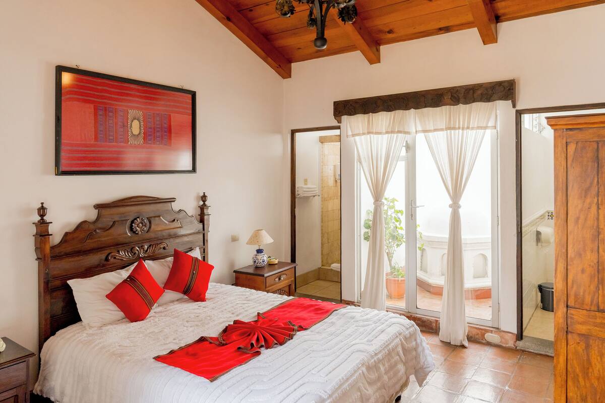 Quarto com cabeceira de madeira, cama com roupa de cama branca e vermelha, mesa de cabeceira com abajur e uma janela grande com cortinas.