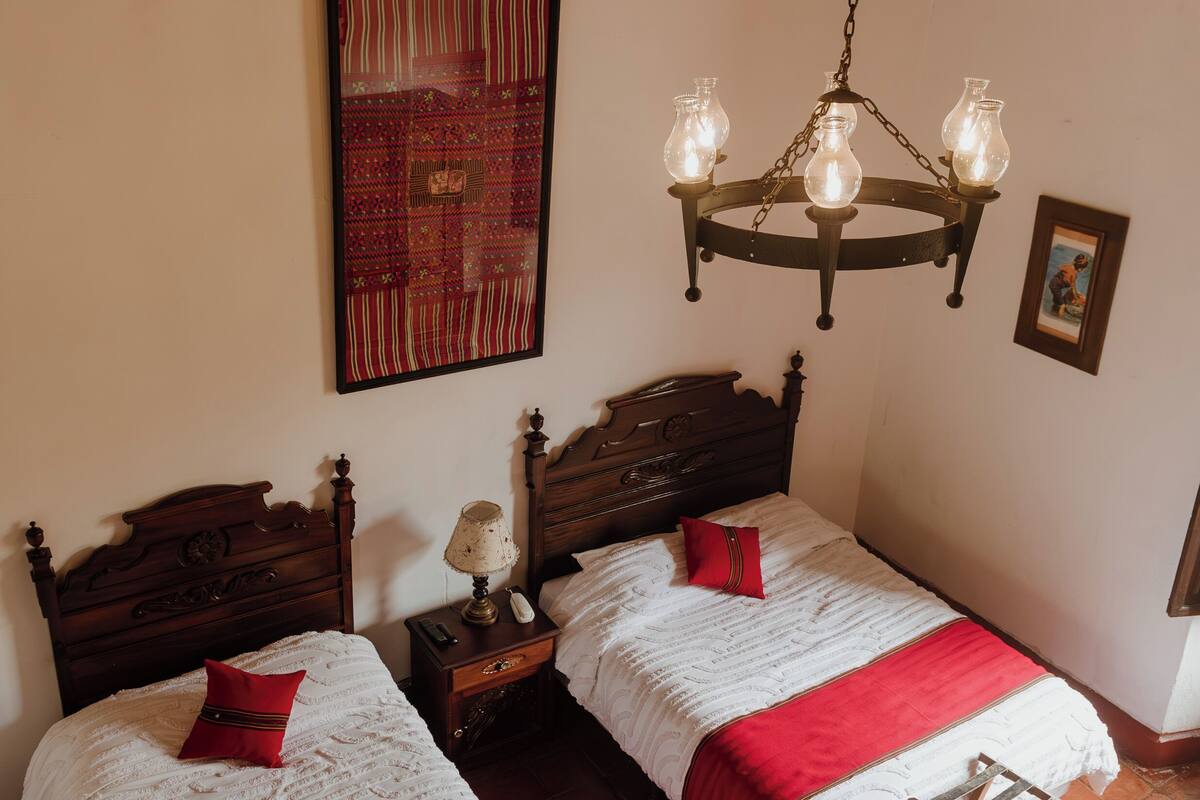 Quarto com duas camas, teto de madeira, luminária pendente e tapeçaria na parede.