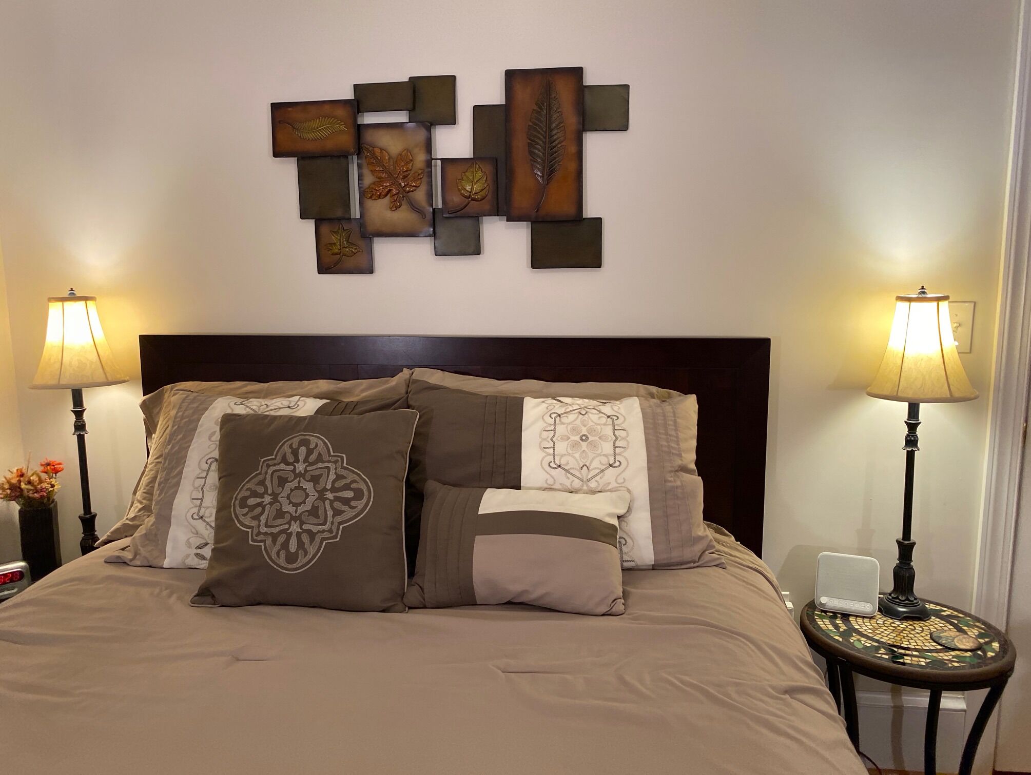 Cama bem arrumada com cabeceira escura, duas luminárias laterais e uma obra de arte na parede acima.