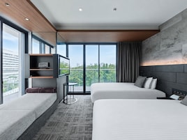 Quarto de hotel moderno com duas camas, uma janela grande com vista para a cidade, uma TV de tela plana e uma mesinha de cabeceira.
