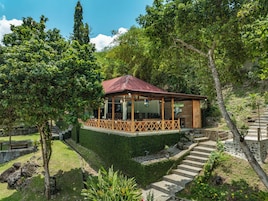 Uma estrutura em estilo gazebo com telhado vermelho, rodeada por vegetação exuberante e degraus de pedra que levam até ela.