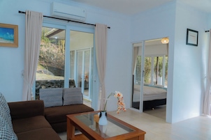Sala de estar com sofá marrom, mesa de centro de vidro e vista para um quarto através de portas de vidro.