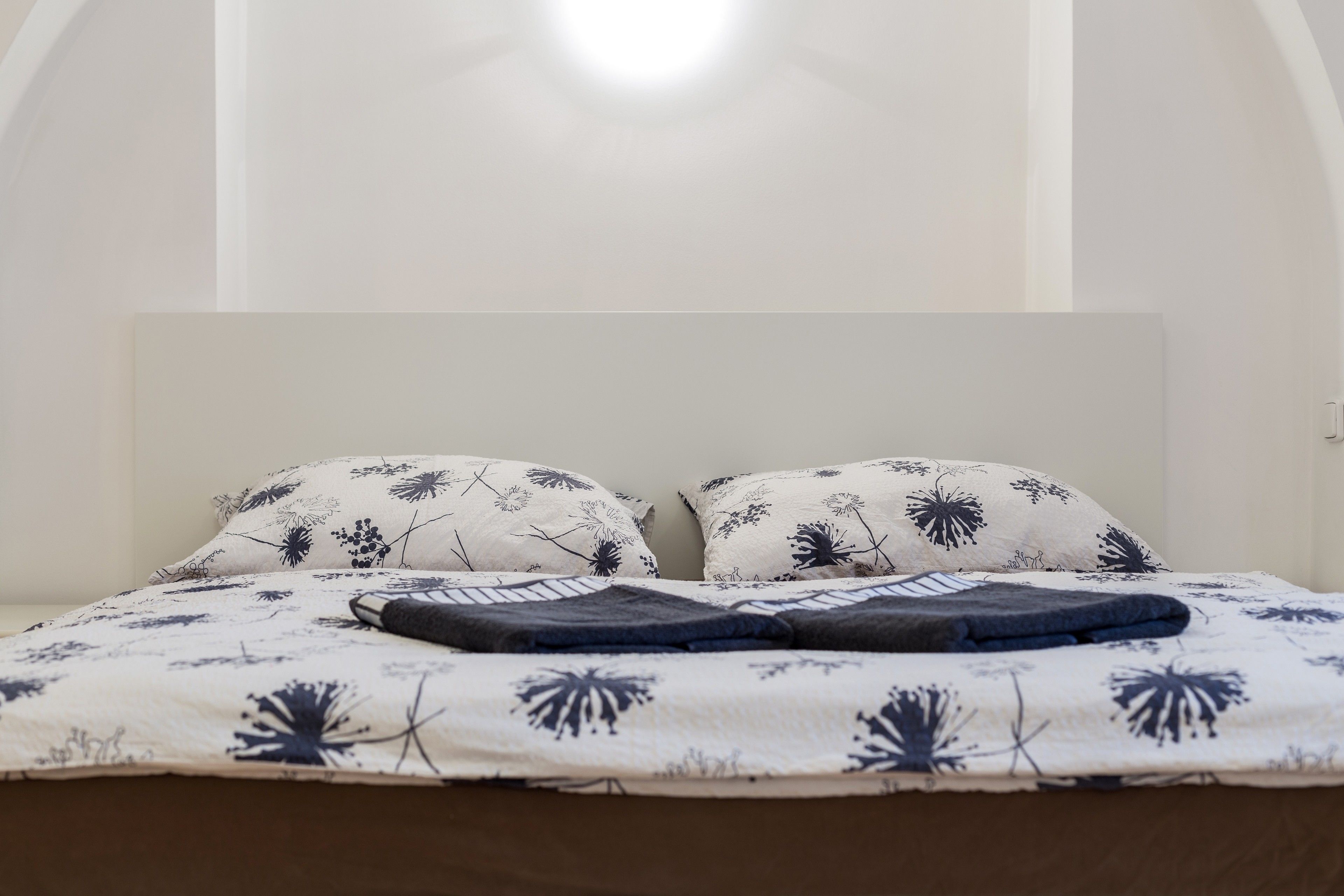 Uma cama com colcha estampada em flor e duas almofadas, cabeceira e parede com luminária.