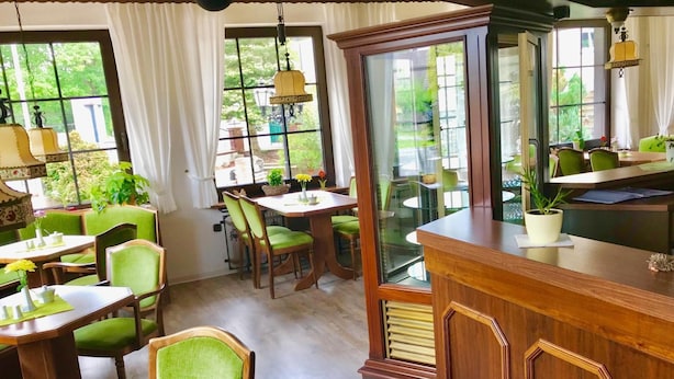 Um restaurante com mobiliário de madeira, cadeiras estofadas em verde e amplas janelas com vista para o verde.