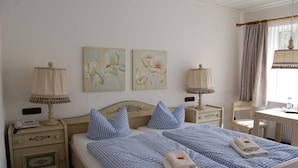 Uma cama de casal com roupa de cama xadrez, duas mesas de cabeceira com abajures, uma escrivaninha pequena e uma cadeira.