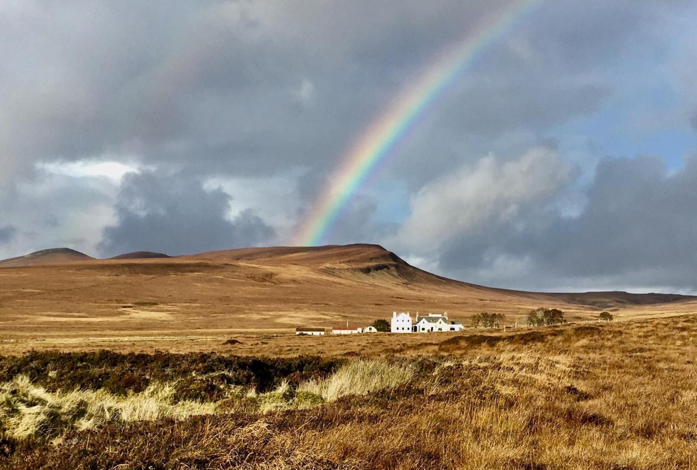 Paisagem rural com colinas ondulantes, uma casa branca e um arco-íris.