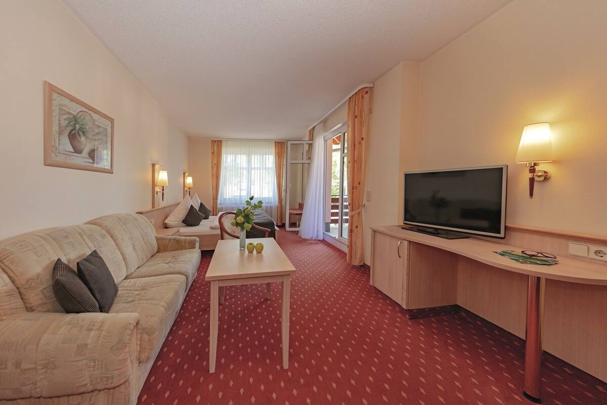 Quarto de hotel com sofá, televisão, uma mesa pequena com um vaso e um tapete vermelho.