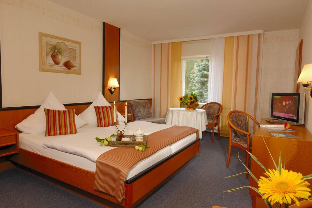 Quarto de hotel com cama, mesa de cabeceira, luminária, televisão e uma pequena mesa com um vaso de flores.
