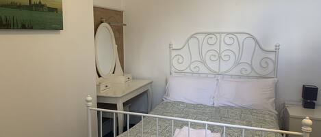 Quarto com estrutura de cama em metal branco, mesa de cabeceira branca, espelho e colcha estampada.