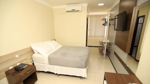 Quarto de hotel com cama, mesa de cabeceira, televisão e pequena escrivaninha.