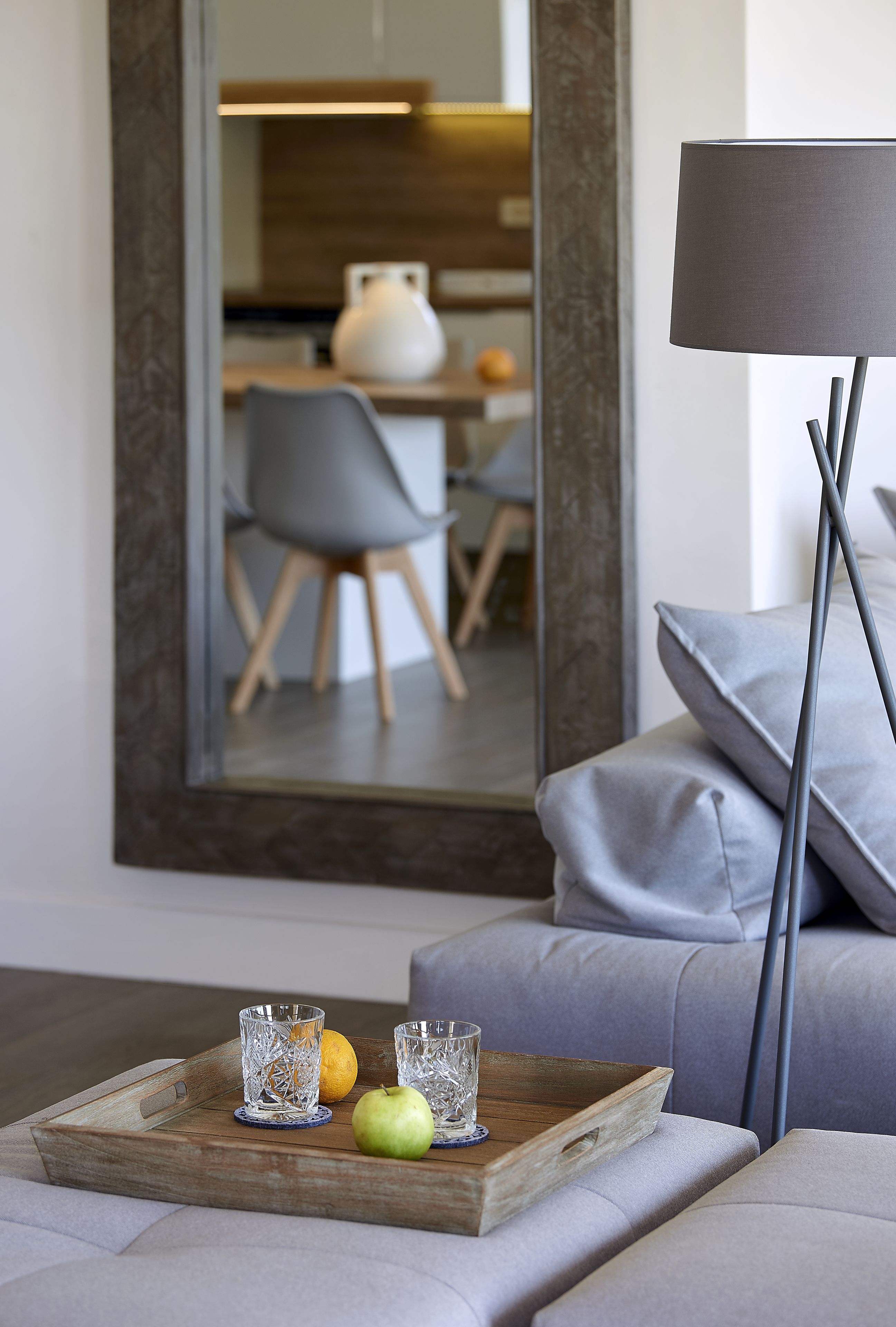 Uma sala de estar moderna com um sofá, uma mesinha lateral com uma bandeja de frutas e copos, um abajur e um espelho grande refletindo o ambiente.