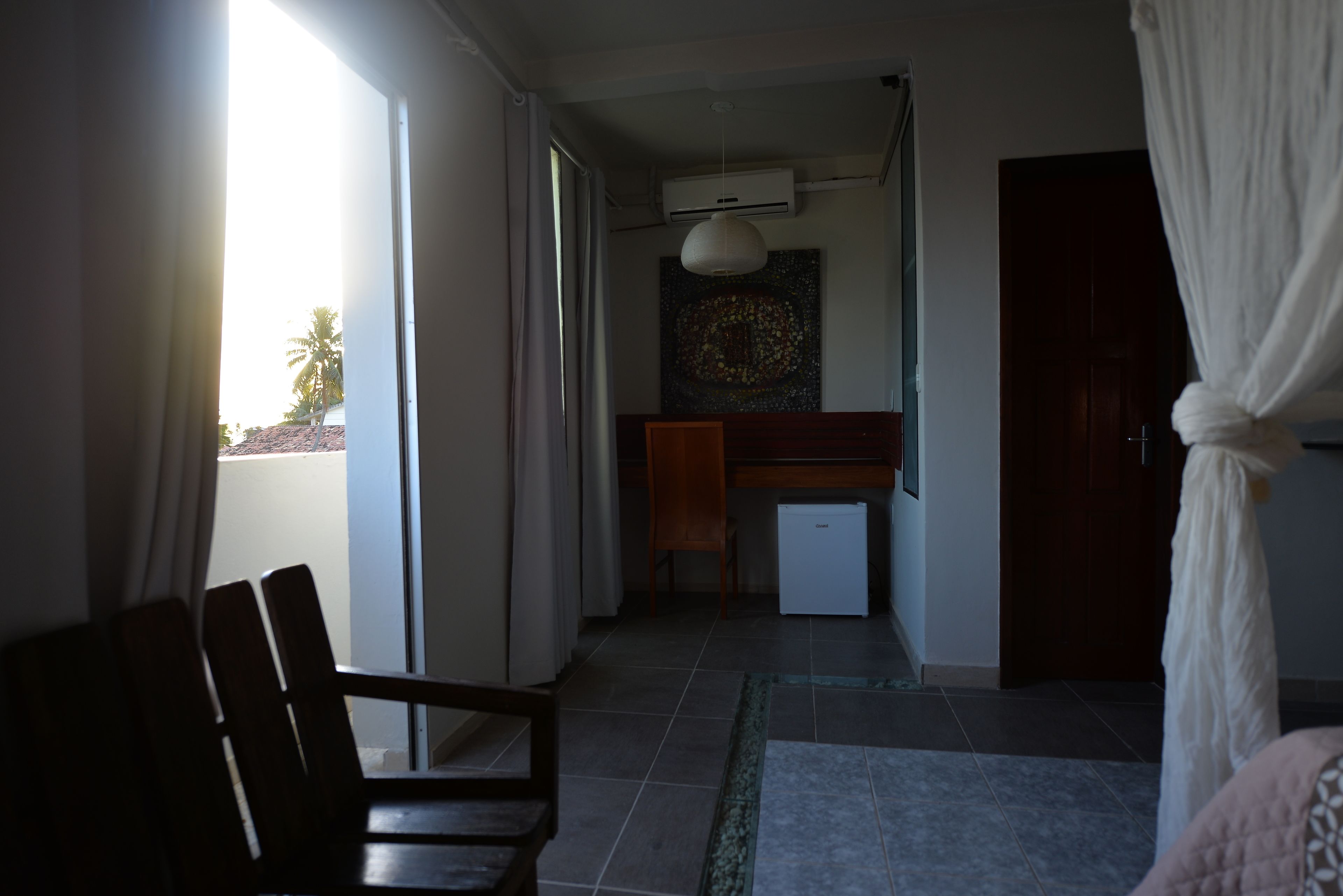 Quarto com cama, escrivaninha e cadeira. Há uma janela com cortinas, um pequeno frigorífico e ar condicionado fixado na parede.