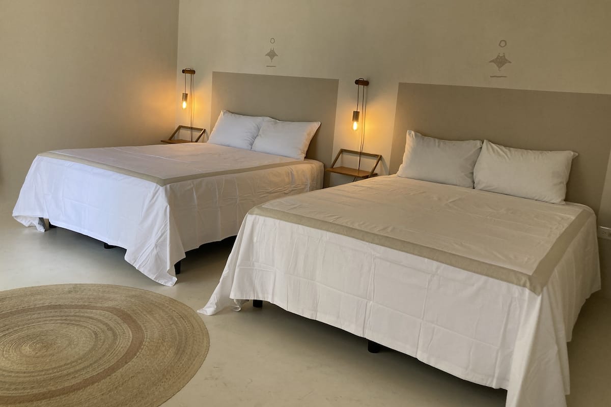 Duas camas de solteiro com roupa de cama branca, um tapete redondo tecido e luminárias fixadas na parede.