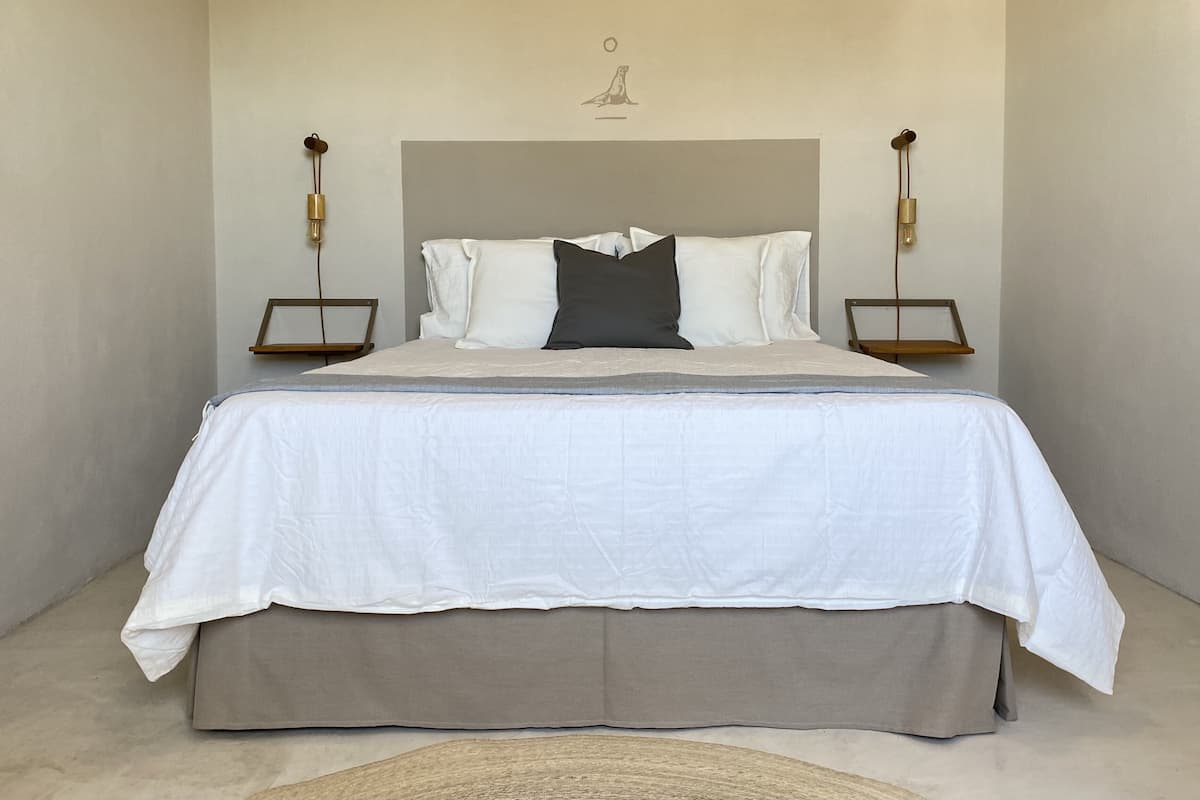 Quarto com cama, almofadas, mesas de cabeceira e luminária fixada na parede.