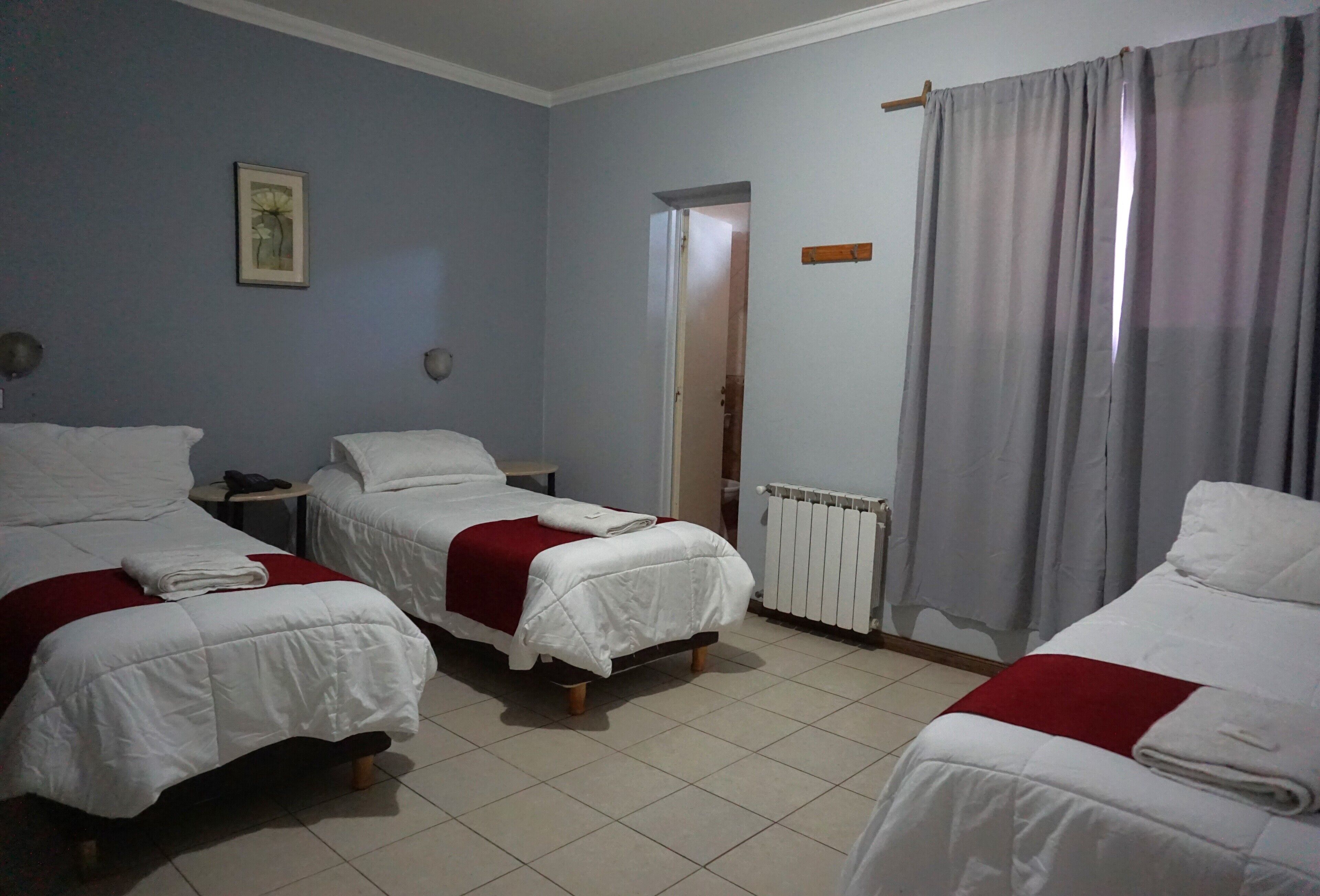 Quarto de hotel com três camas, uma mesinha e um radiador.