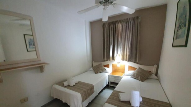 Quarto de hotel com duas camas de solteiro, um ventilador de teto, mesa de cabeceira com abajur e janela com cortinas.