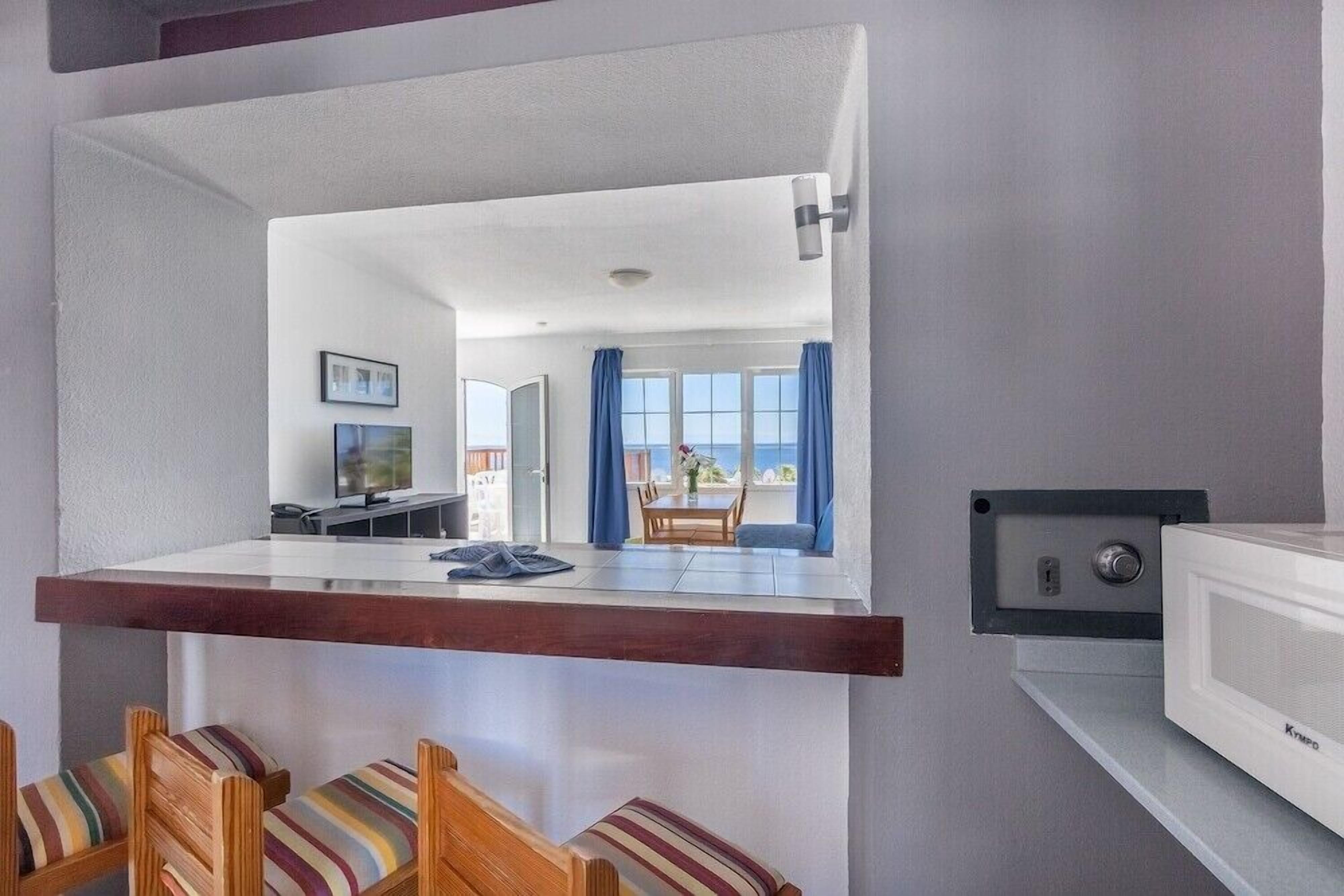 Uma cozinha compacta com bancada de madeira, micro-ondas e vista para a área de refeições através de uma grande janela com cortinas azuis.
