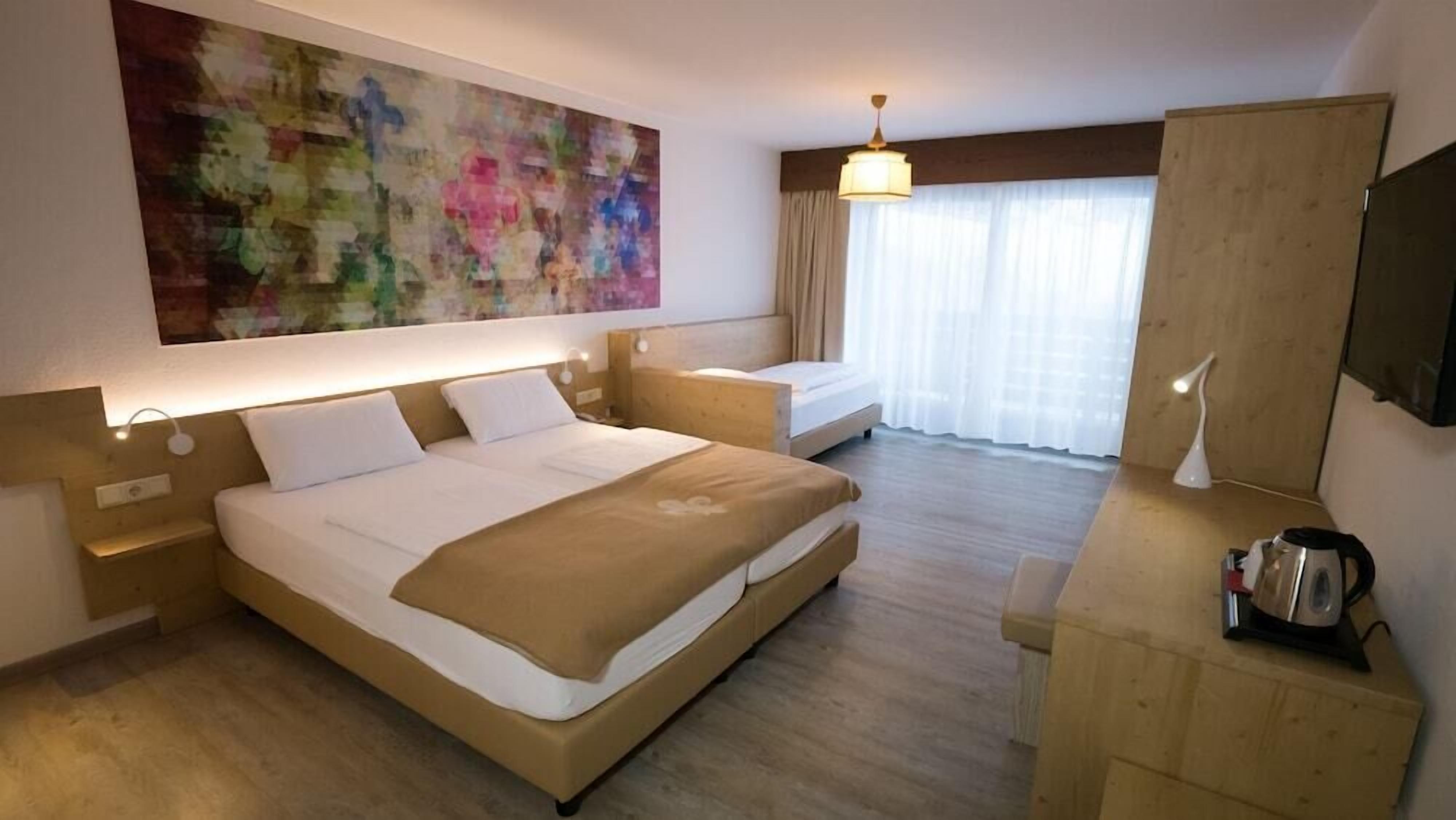 Quarto de hotel com uma cama grande, uma cama menor, uma escrivaninha com chaleira, um quadro abstrato de grandes dimensões e uma janela com cortinas.