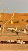 Paisagem desértica com área de refeições, com cadeiras e mesas brancas, uma árvore com galhos secos e uma luminária decorativa pendente.