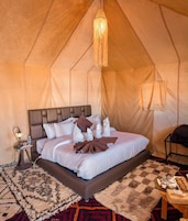 Um quarto tipo tenda com uma cama, uma luminária pendente, uma mesinha de cabeceira com abajur e uma pequena mesa com uma bandeja de objetos.