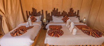 Quarto de hotel com duas camas, cada uma com uma toalha e um edredom estampado. Há duas mesas de cabeceira com luminárias e uma garrafa de água.