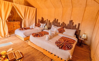 Quarto com duas camas, cabeceira de madeira, tapete tecido e mesa de cabeceira de madeira com abajur.