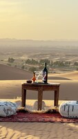 Uma mesa com uma garrafa e copos sobre um tapete de palha, com dunas de areia e colinas ao fundo.