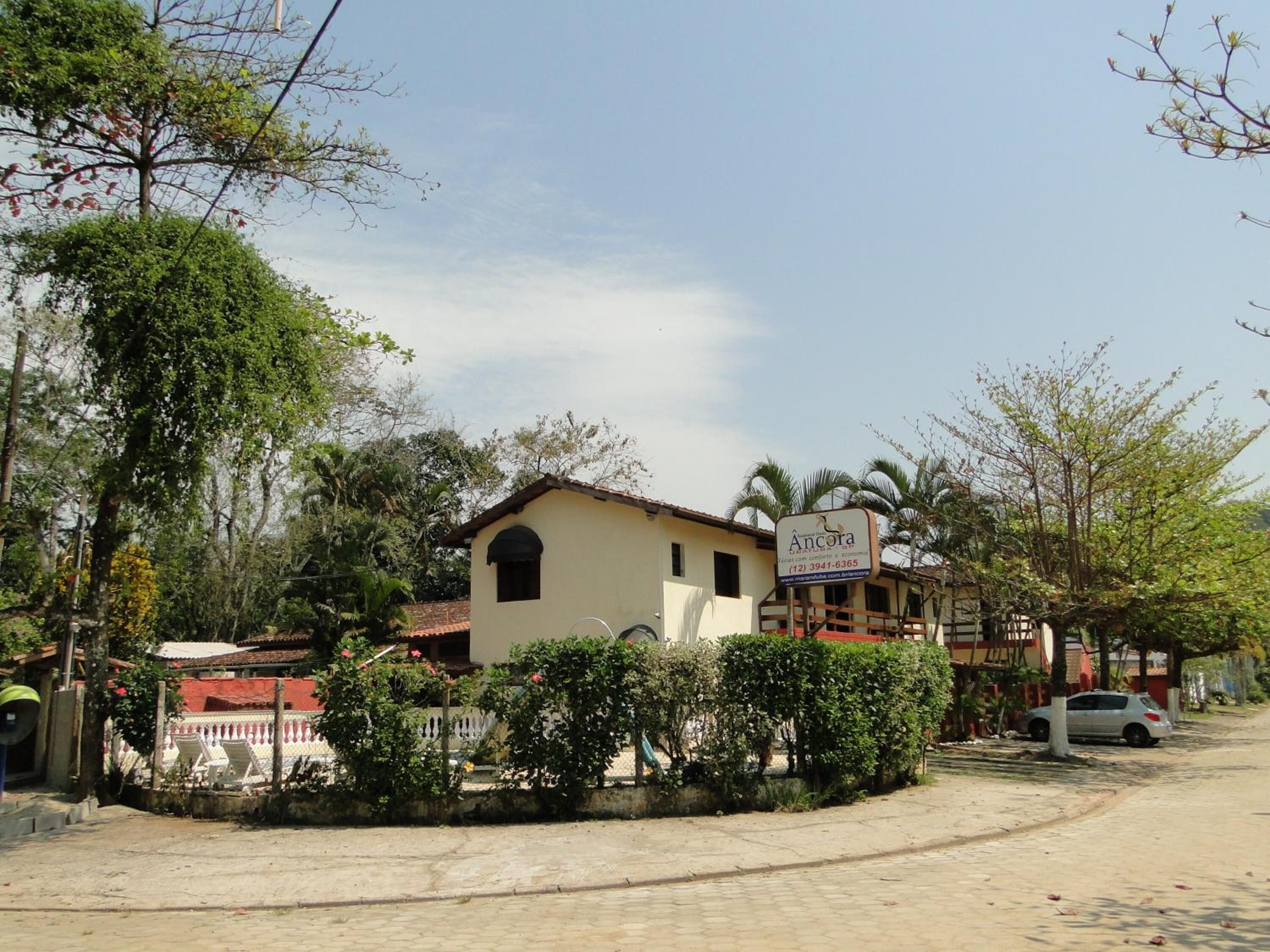 Um edifício de dois andares com fachada bege e telhado vermelho, rodeado por vegetação e uma rua pavimentada.