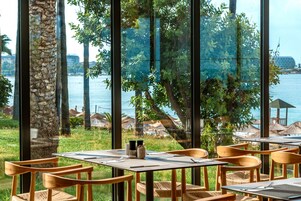 Uma área de refeições ao ar livre com mesas e cadeiras, janelas amplas e vista para o mar e a paisagem urbana.