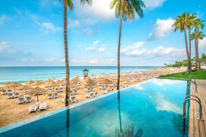 Vista da piscina com praia, cadeiras de sol e palmeiras.